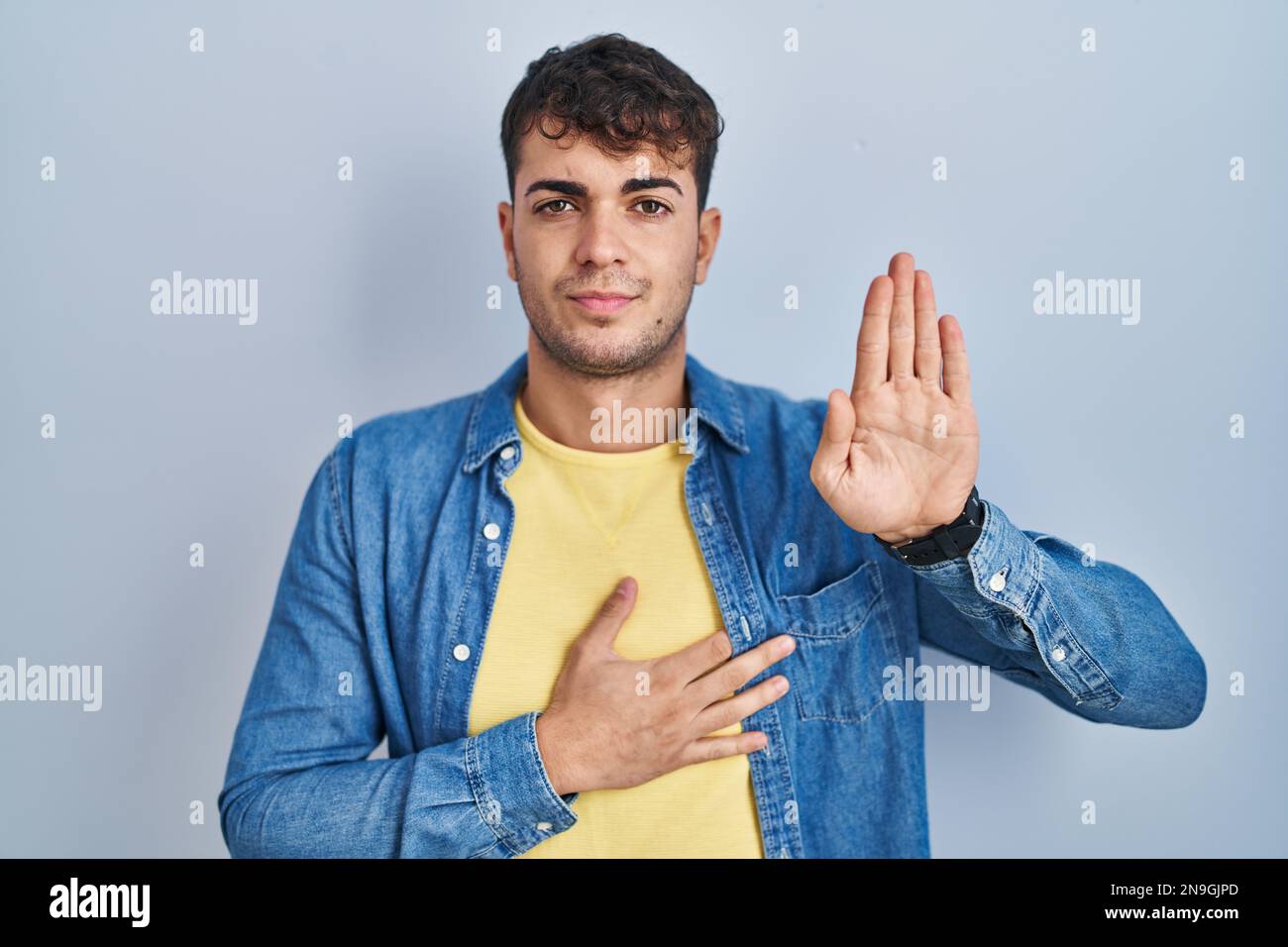 Young hispanic man standing over blue background swearing with hand on ...