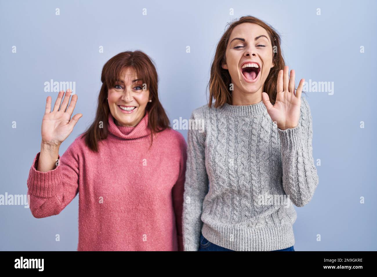 Mother and daughter standing over blue background waiving saying hello ...