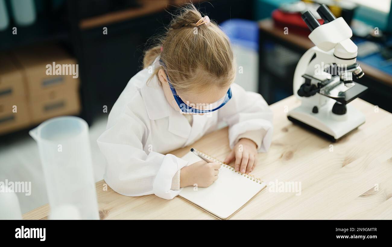 Adorable blonde girl student using microscope writing on notebook at ...