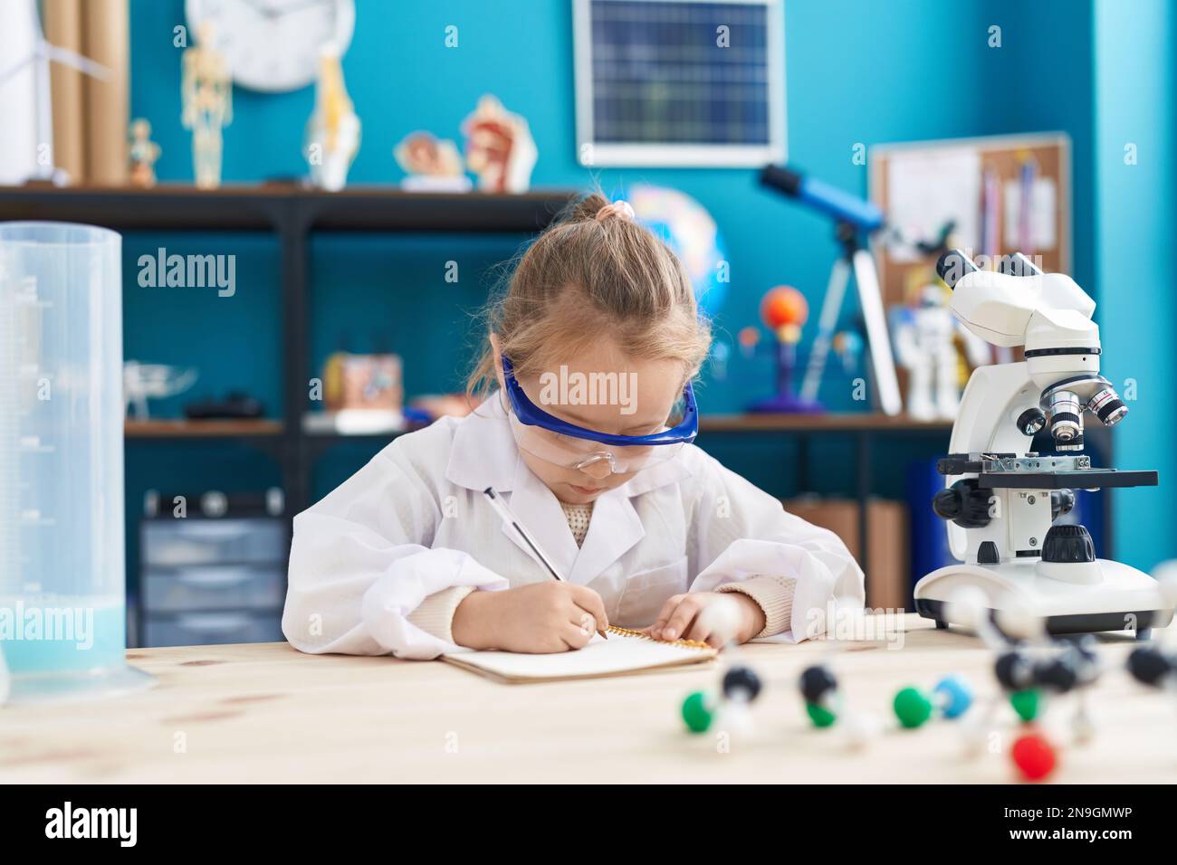 Adorable blonde girl student using microscope writing on notebook at ...