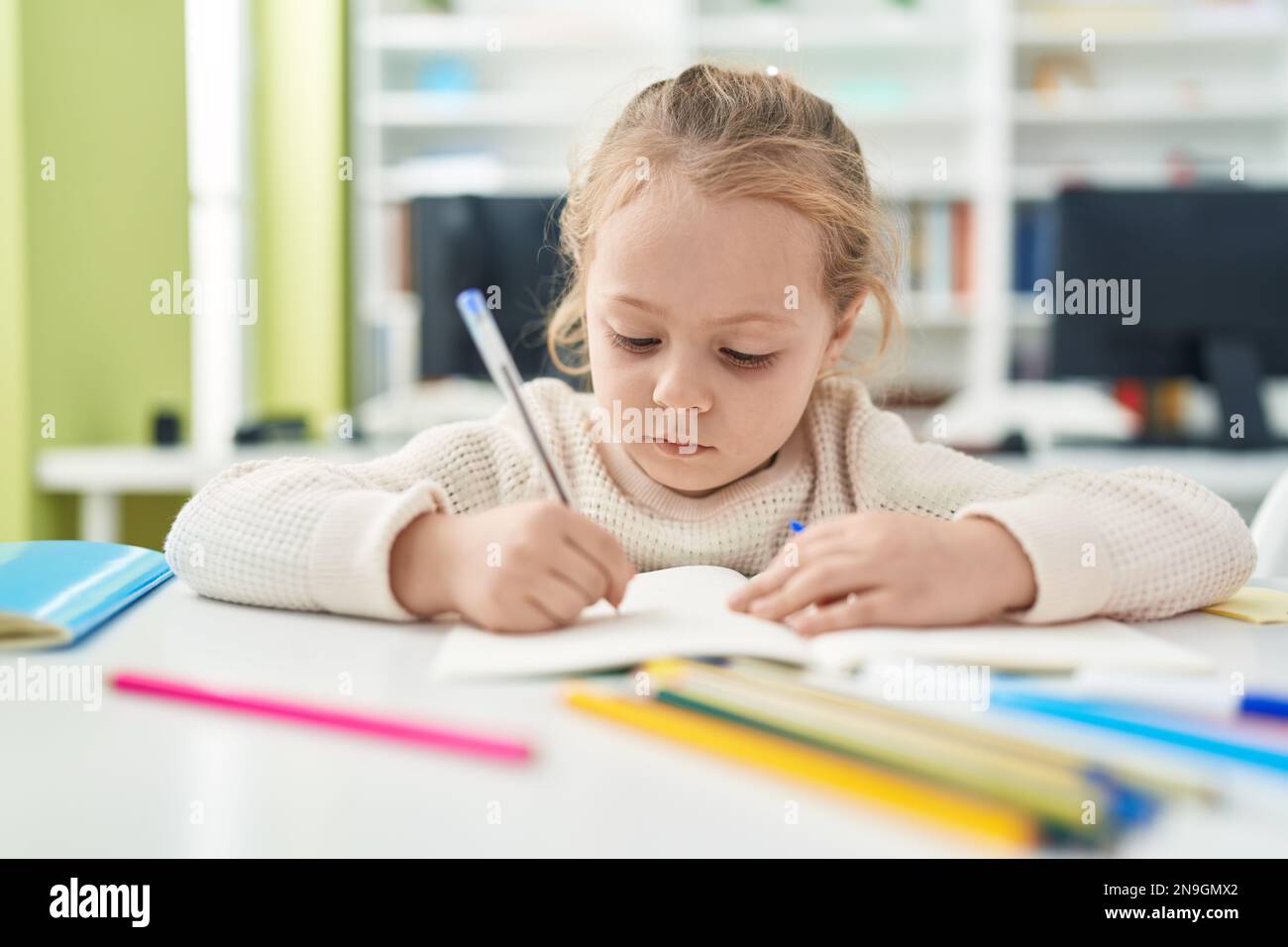 Adorable blonde girl student writing on notebook sitting on table at ...