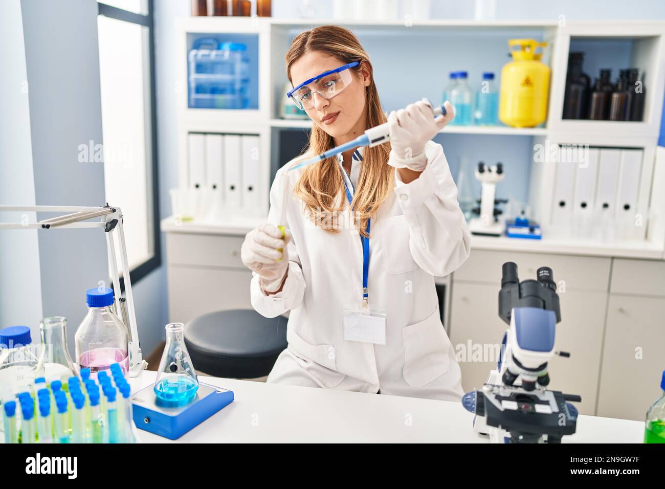 Young blonde woman wearing scientist uniform pouring liquid using ...