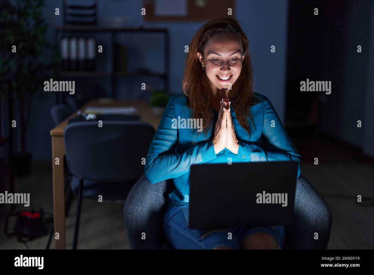 Brunette woman working at the office at night praying with hands ...