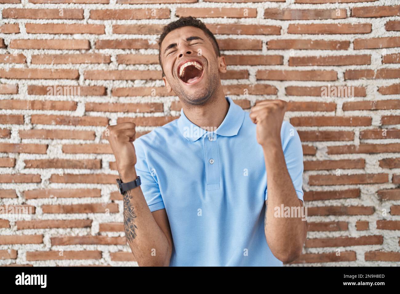 Brazilian young man standing over brick wall celebrating surprised and ...