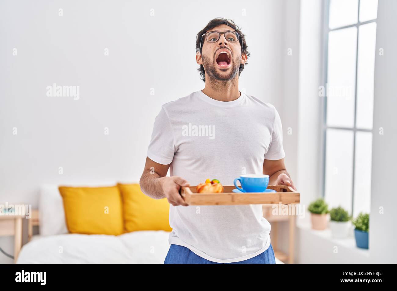Handsome latin man eating breakfast on the bed angry and mad screaming ...