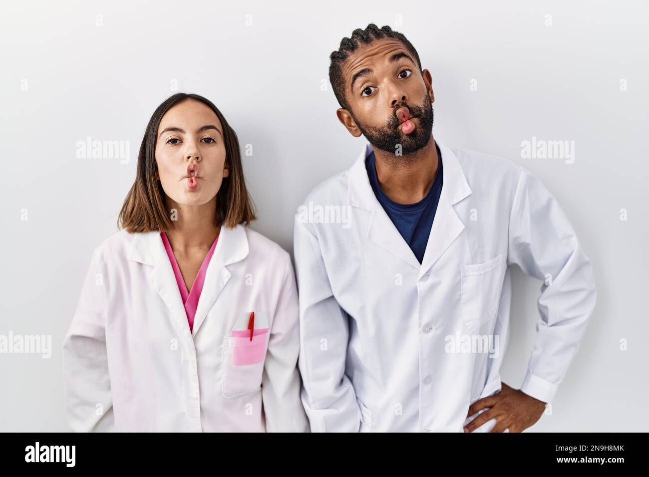 Young hispanic doctors standing over white background making fish face ...