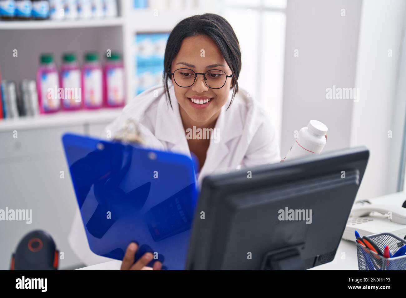 Young hispanic woman pharmacist reading document using computer at ...
