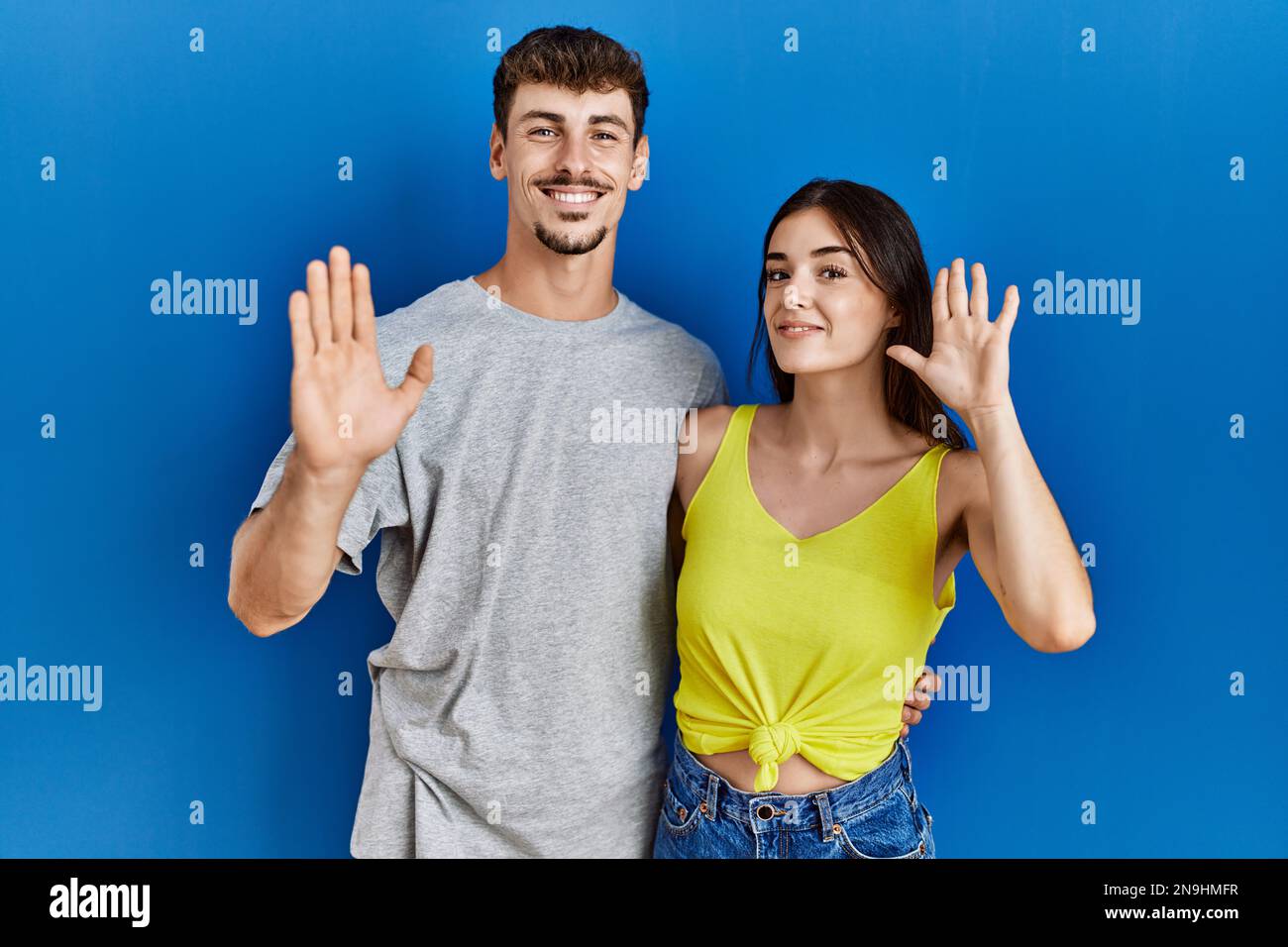 Young hispanic couple standing together over blue background waiving ...
