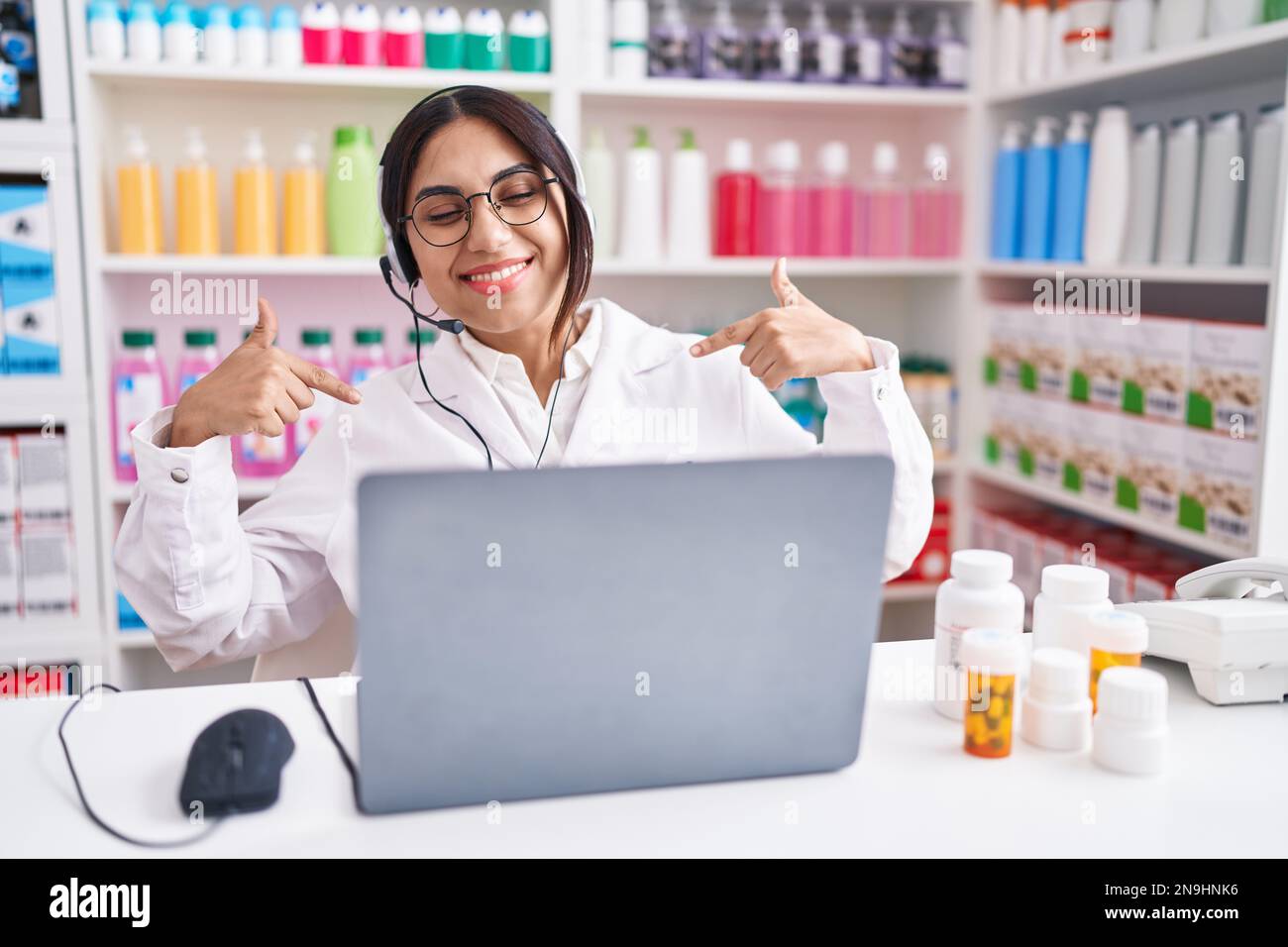 Young arab woman working at pharmacy drugstore using laptop looking confident with smile on face ...