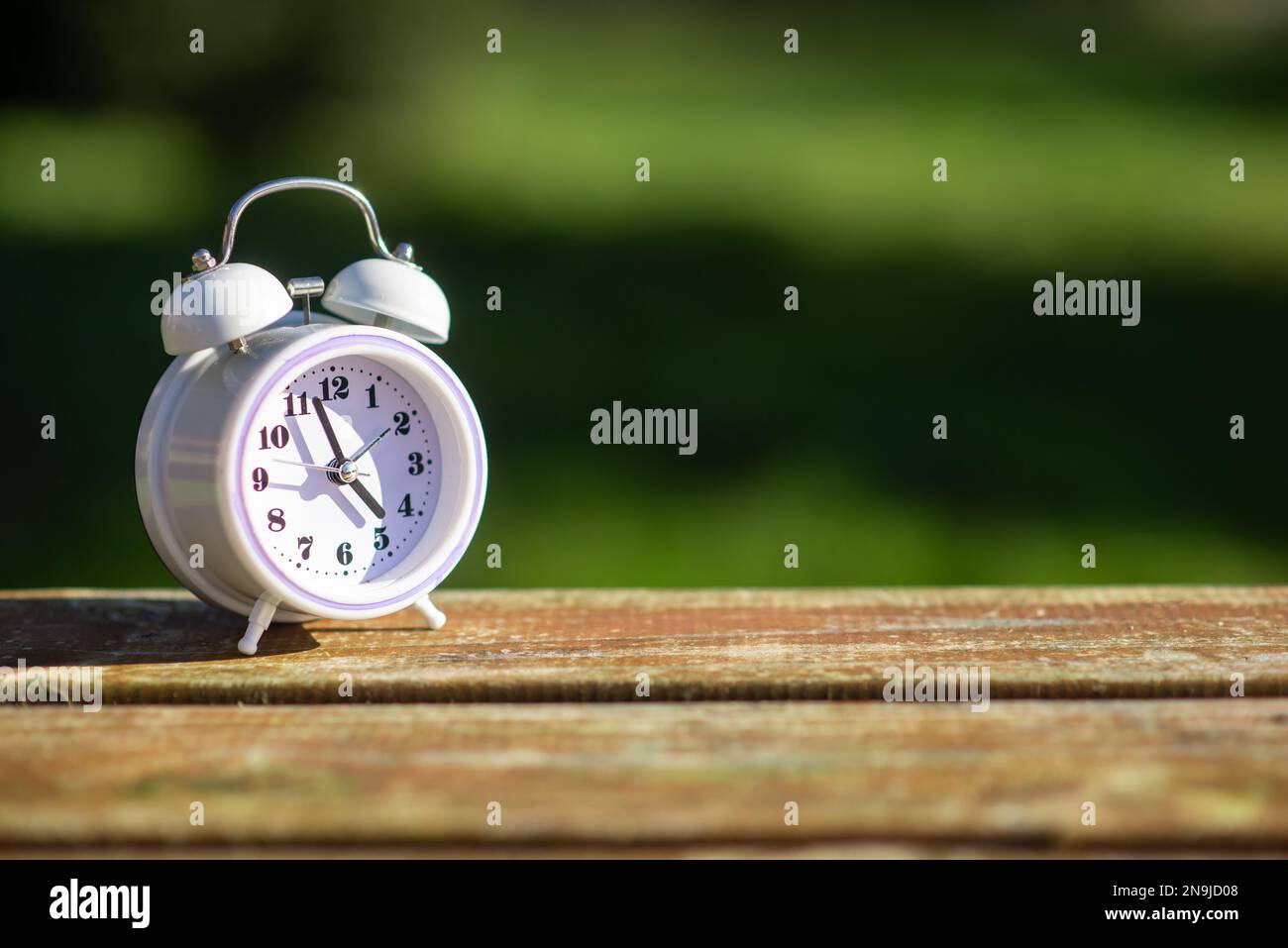 White color alarm clock on a wooden table. Blurred green foliage ...