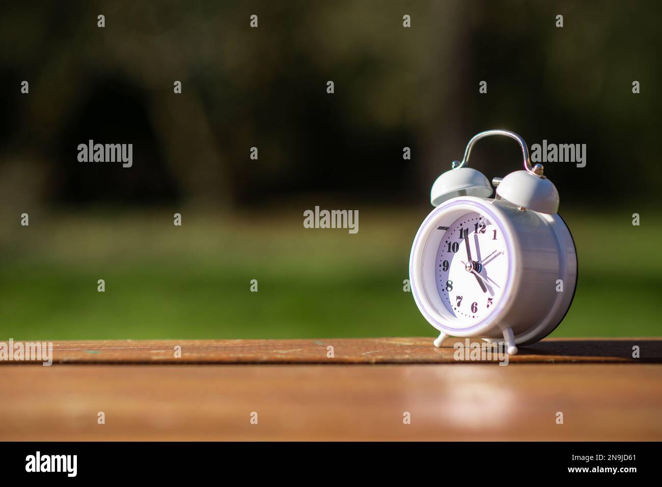White color alarm clock on a wooden table. Blurred green foliage ...