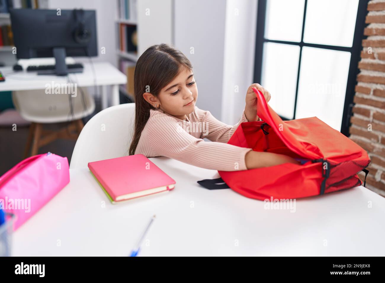 Adorable hispanic girl student sitting on table putting book on ...
