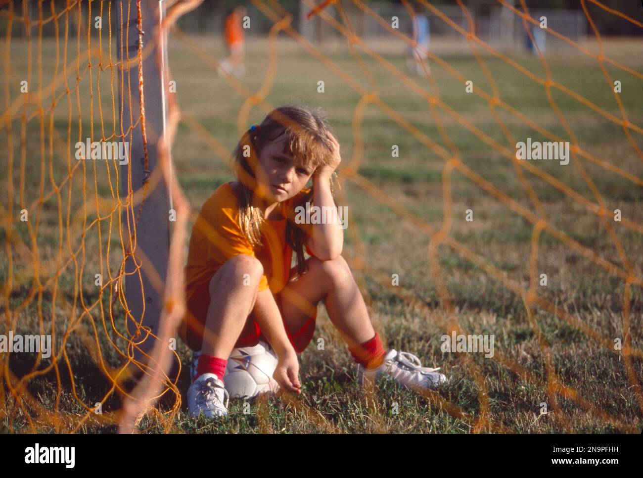 Little girl sitting on a soccer ball. Looking through the goalie net ...