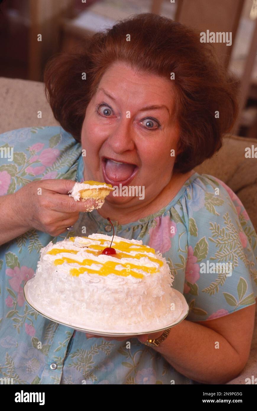 Older overweight woman eating a piece of cake while holding the cake ...