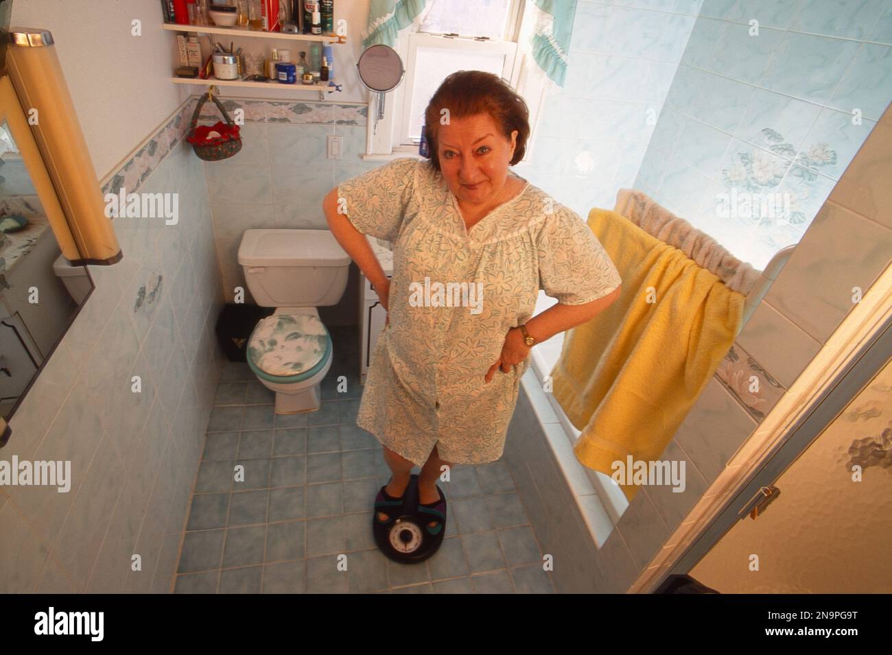 Older Overweight Woman Standing On A Bathroom Scale Looking Up Stock