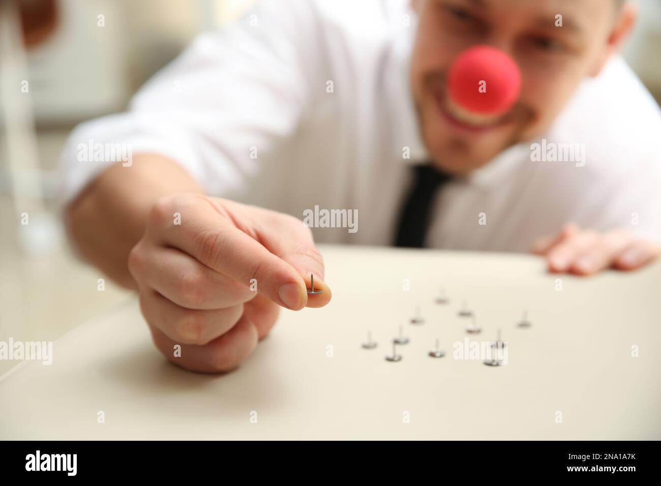 Man with clown nose putting pins onto colleague's chair in office ...