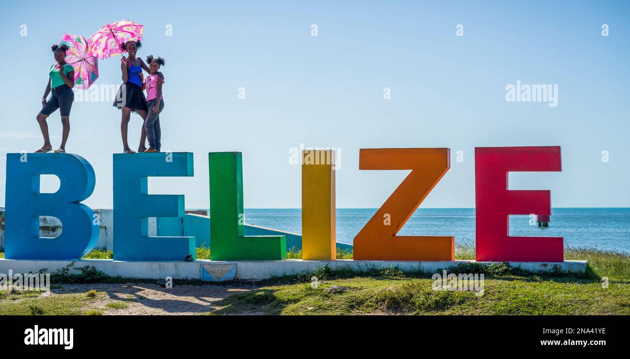 Three girls stand on the colourful Belize sign with a view of the ocean ...