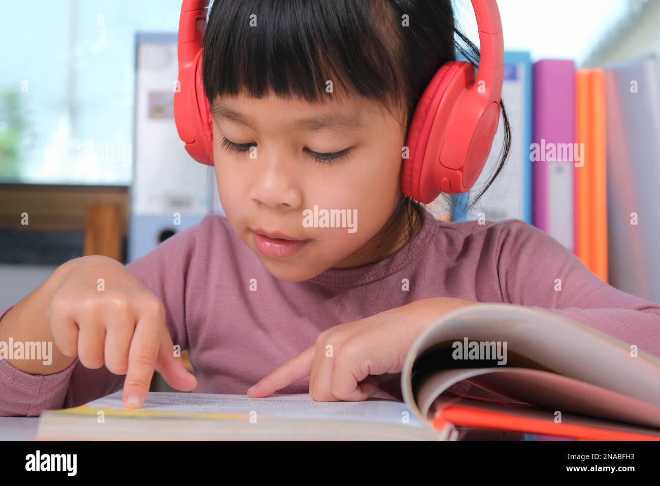 Cheerful little girl in headphones reading a book sits at the table in ...