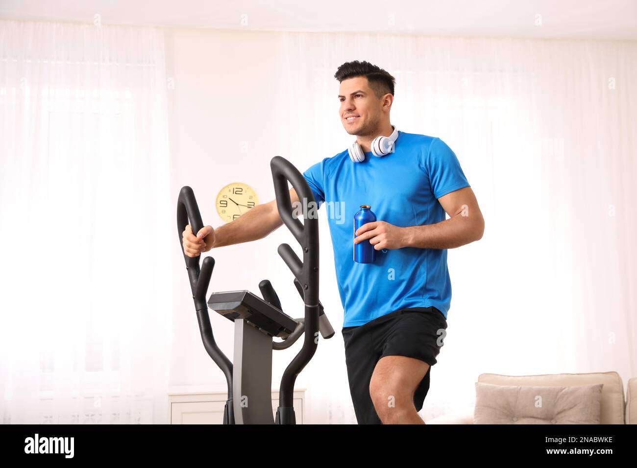 Man with bottle of water using modern elliptical machine at home Stock ...