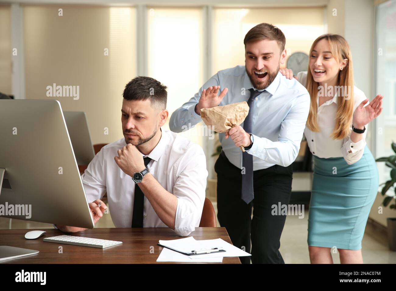 Coworkers popping paper bag behind their colleague in office. Funny ...