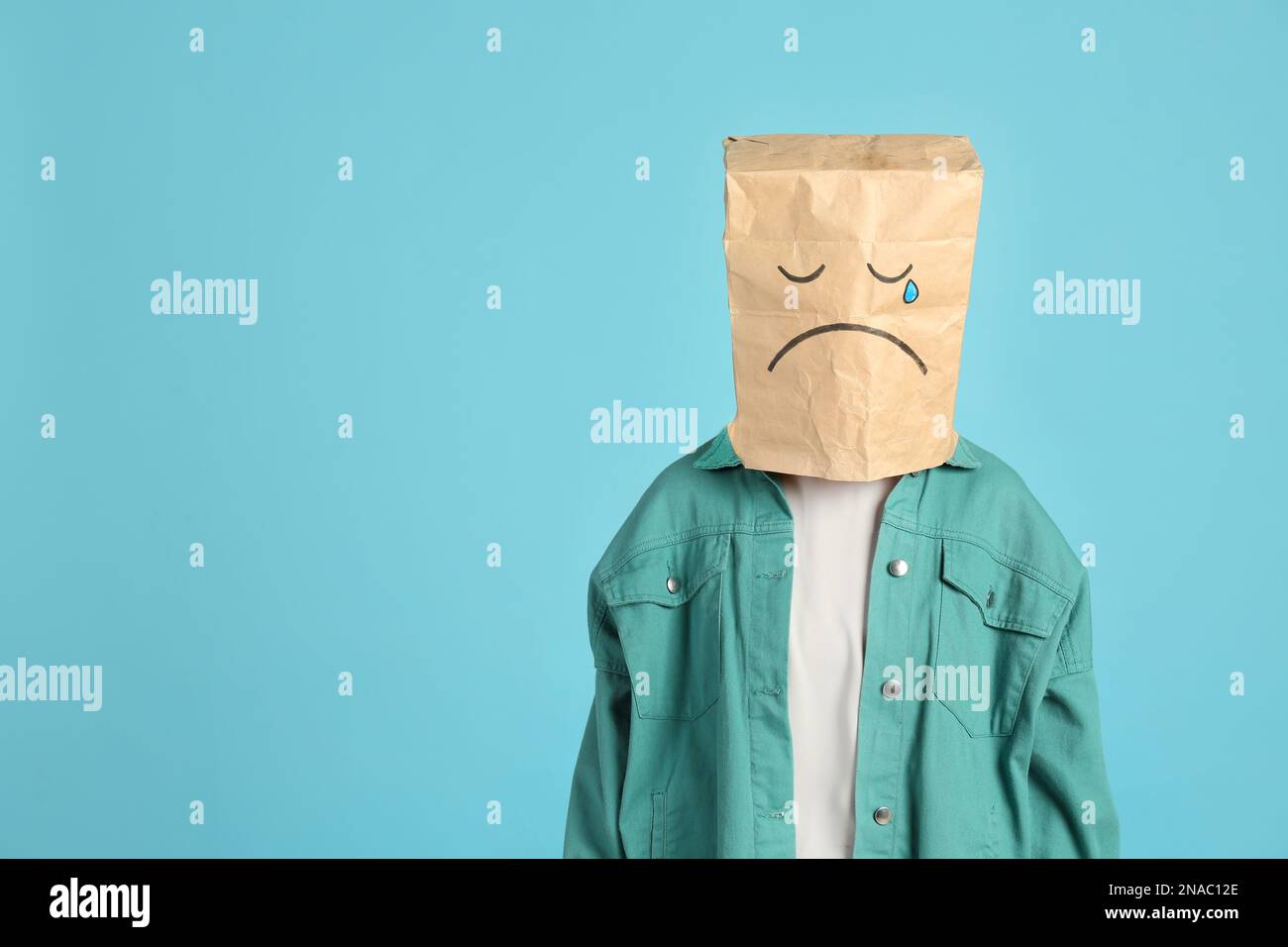 Woman wearing paper bag with drawn sad face on light blue background ...