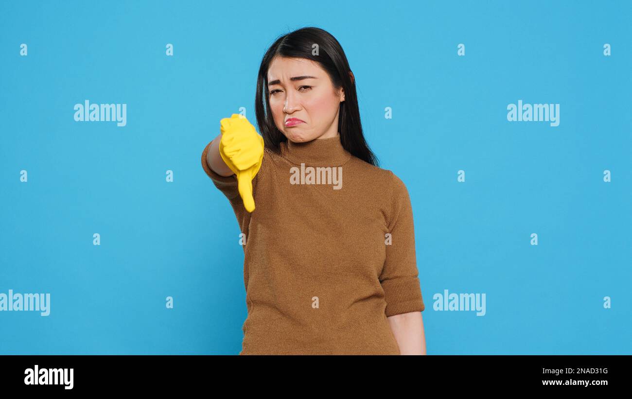 Upset asian maid doing thumbs down gesture while cleaning client house ...