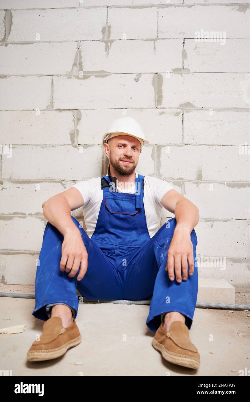 Male builder in work overalls sitting on the floor in building under ...