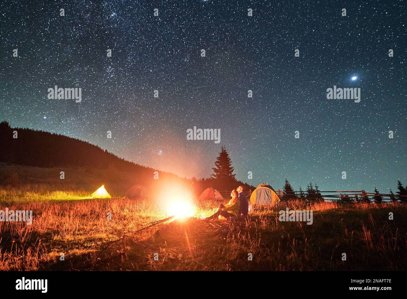 Night camping in mountains under starry sky. Two people, couple sitting on grass near burning ...