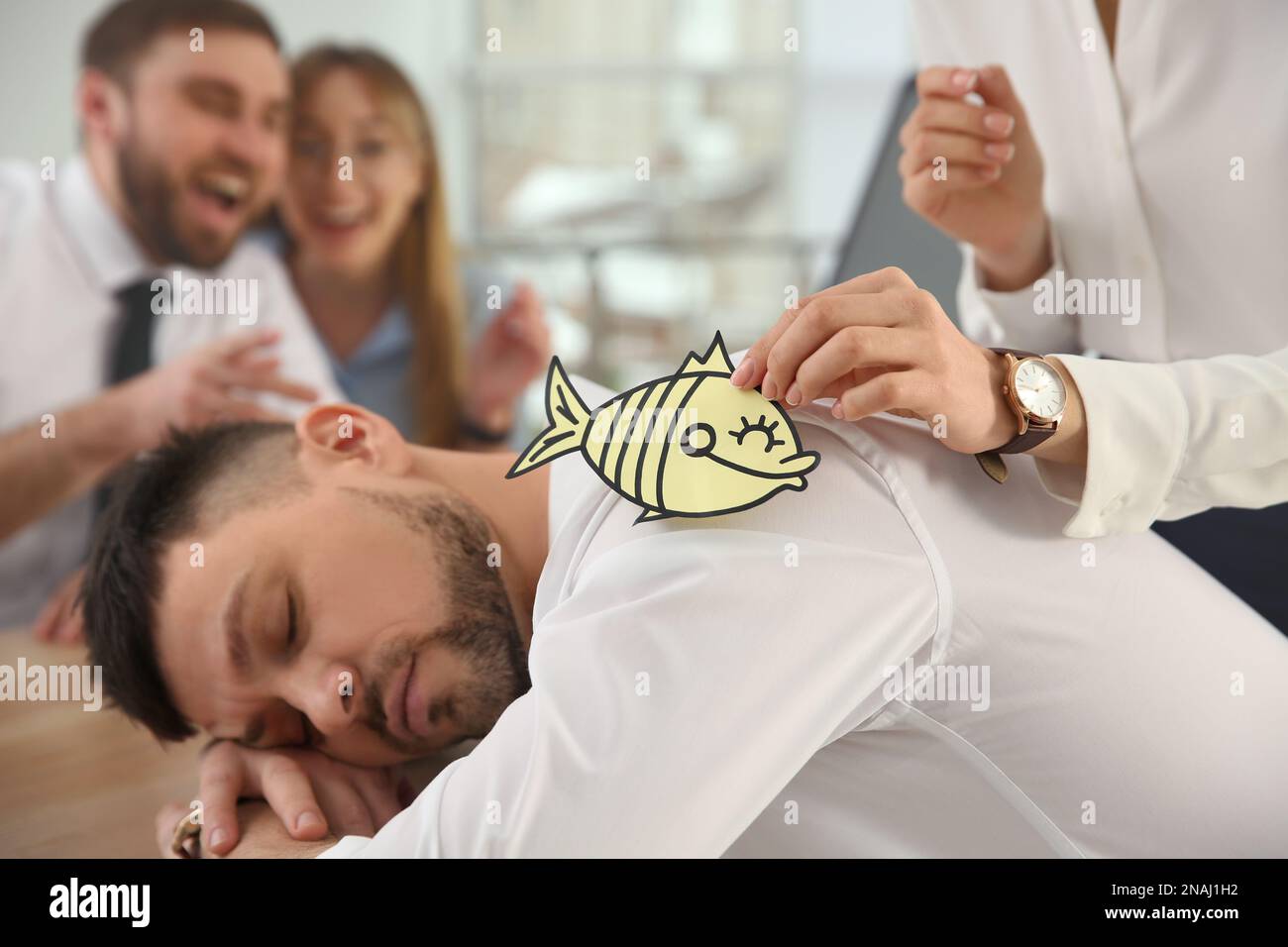 Young woman sticking paper fish to colleague's back while he sleeping ...