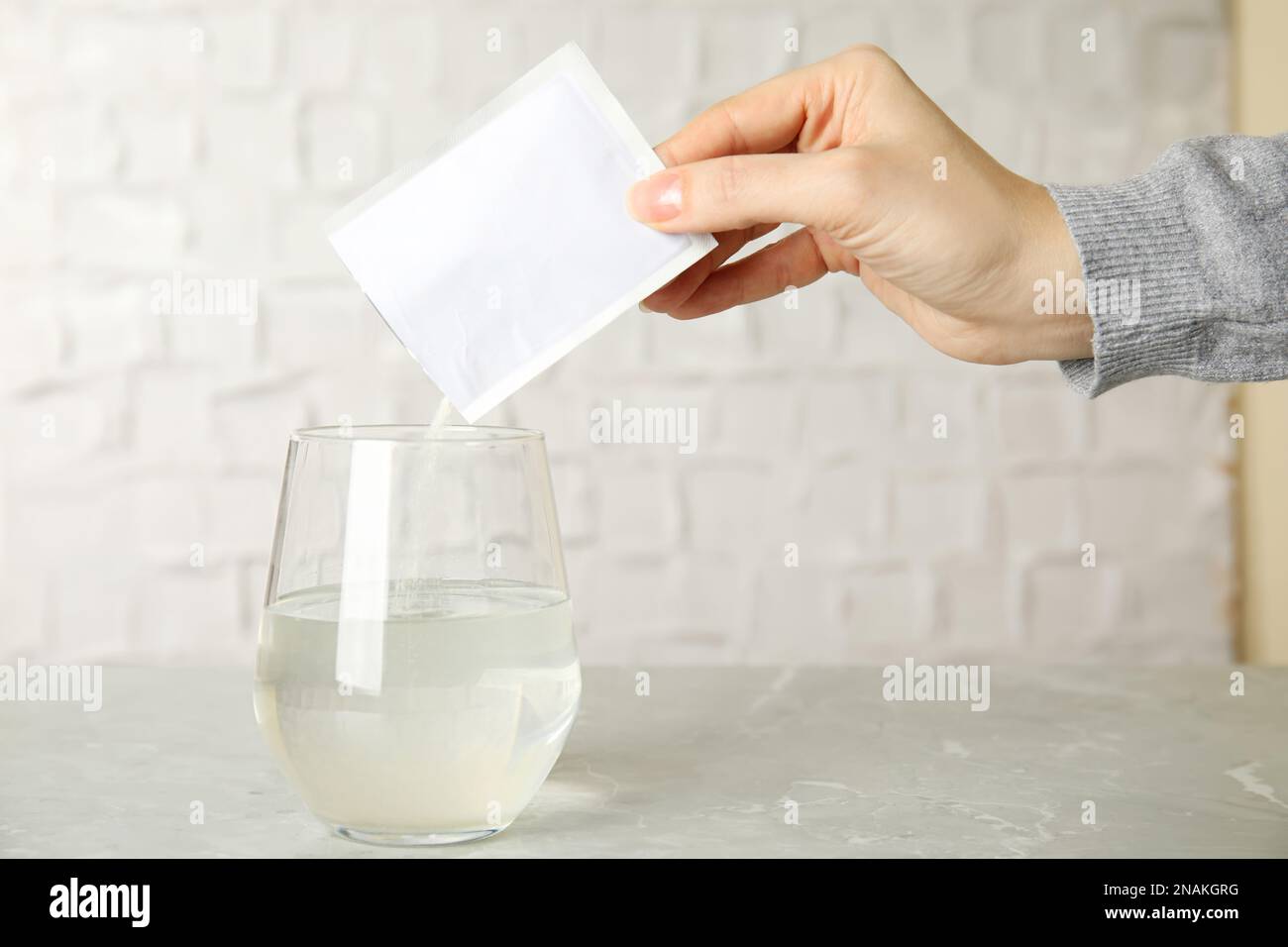 Woman pouring powder from medicine sachet into glass of water on grey ...