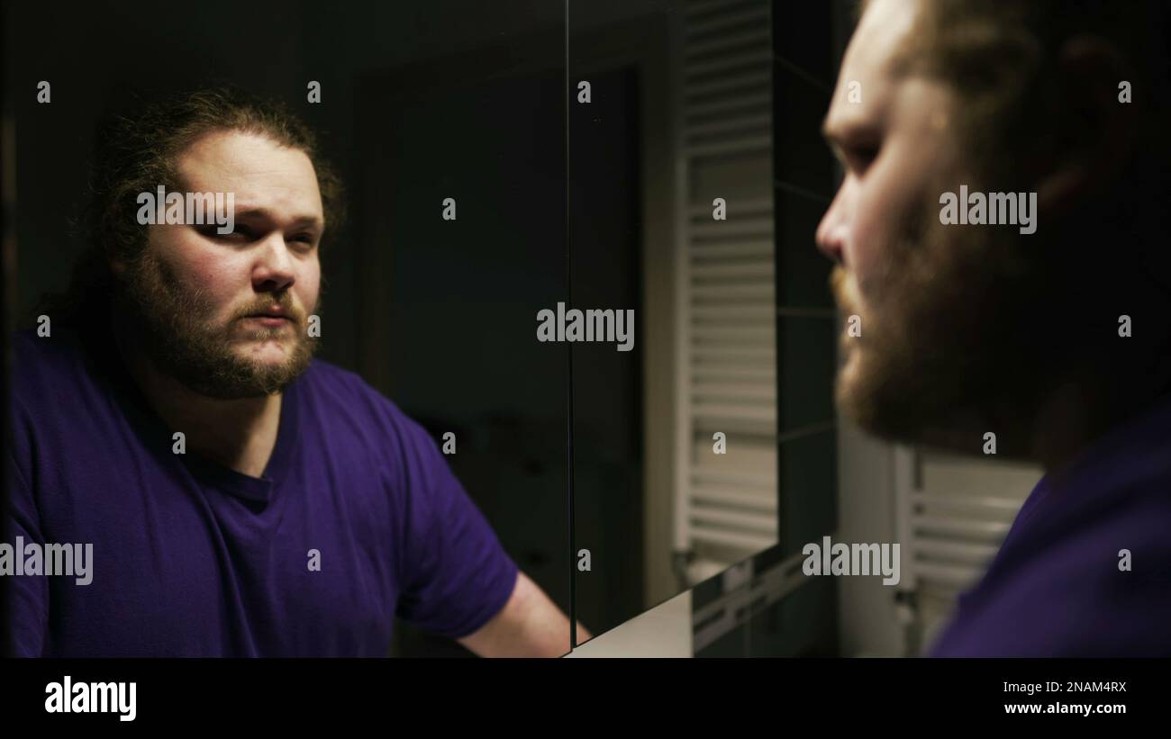 Depressed man staring at own reflection in front of bathroom mirror ...
