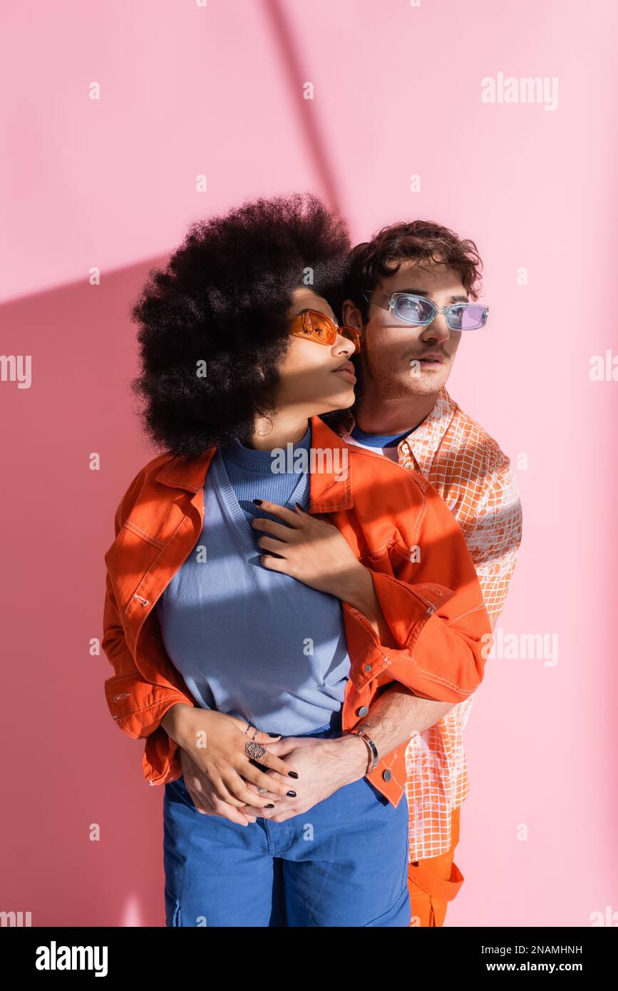 Trendy man in sunglasses hugging african american woman on pink ...