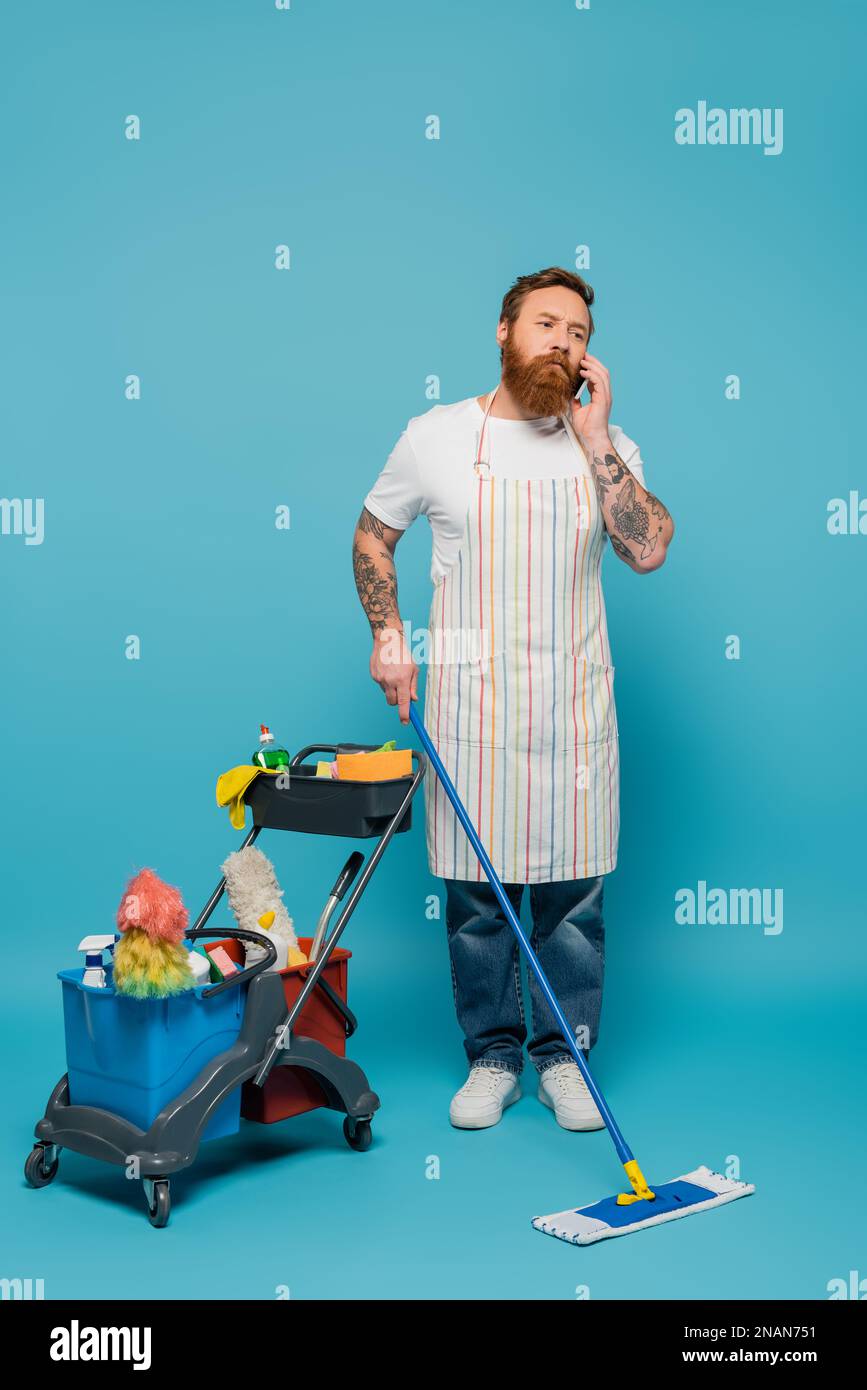serious man with mop talking on mobile phone near cart with cleaning ...