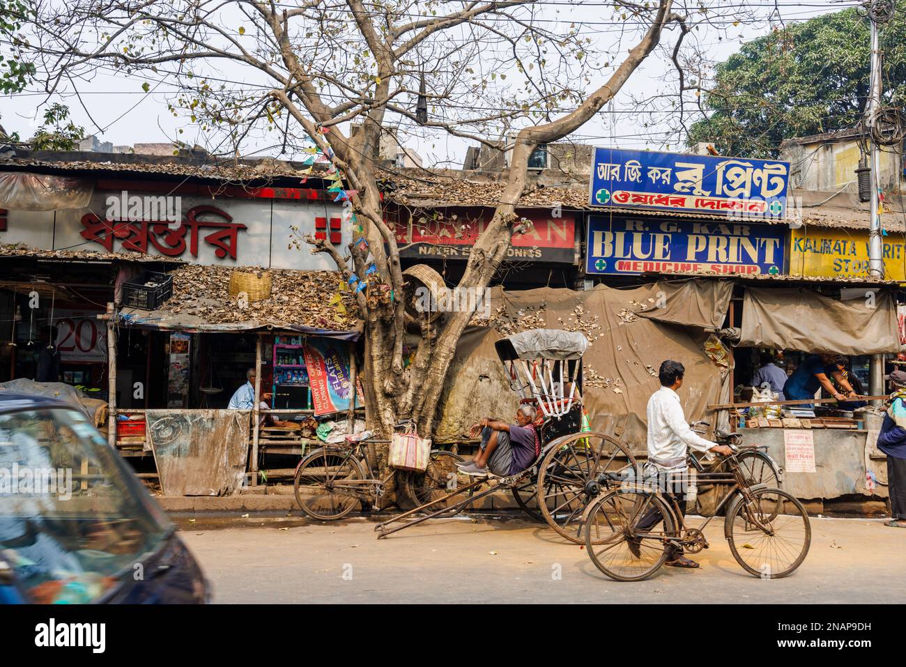 Street scene of a resting rickshaw driver, local people, shops, stalls ...