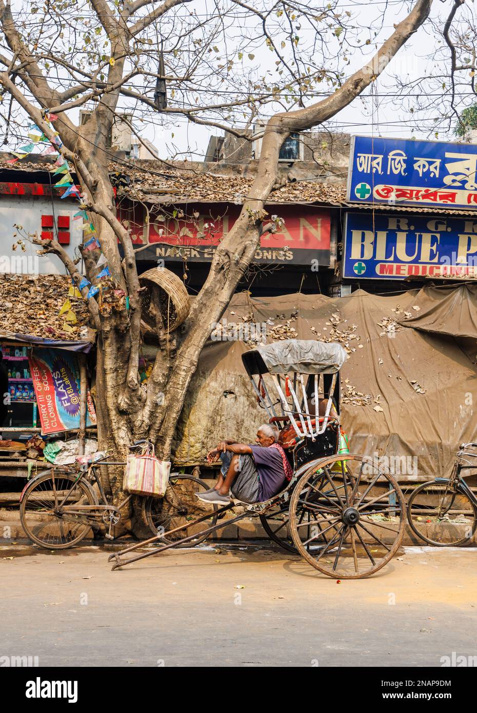 Street scene of a resting rickshaw driver, local people, shops, stalls ...