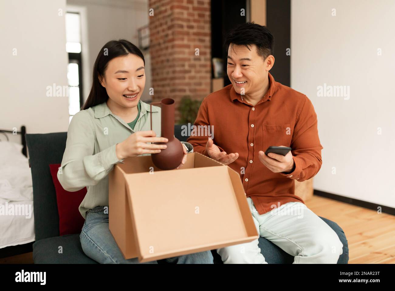 Happy asian couple unpacking box with vase after successful shopping and delivery, sitting on ...