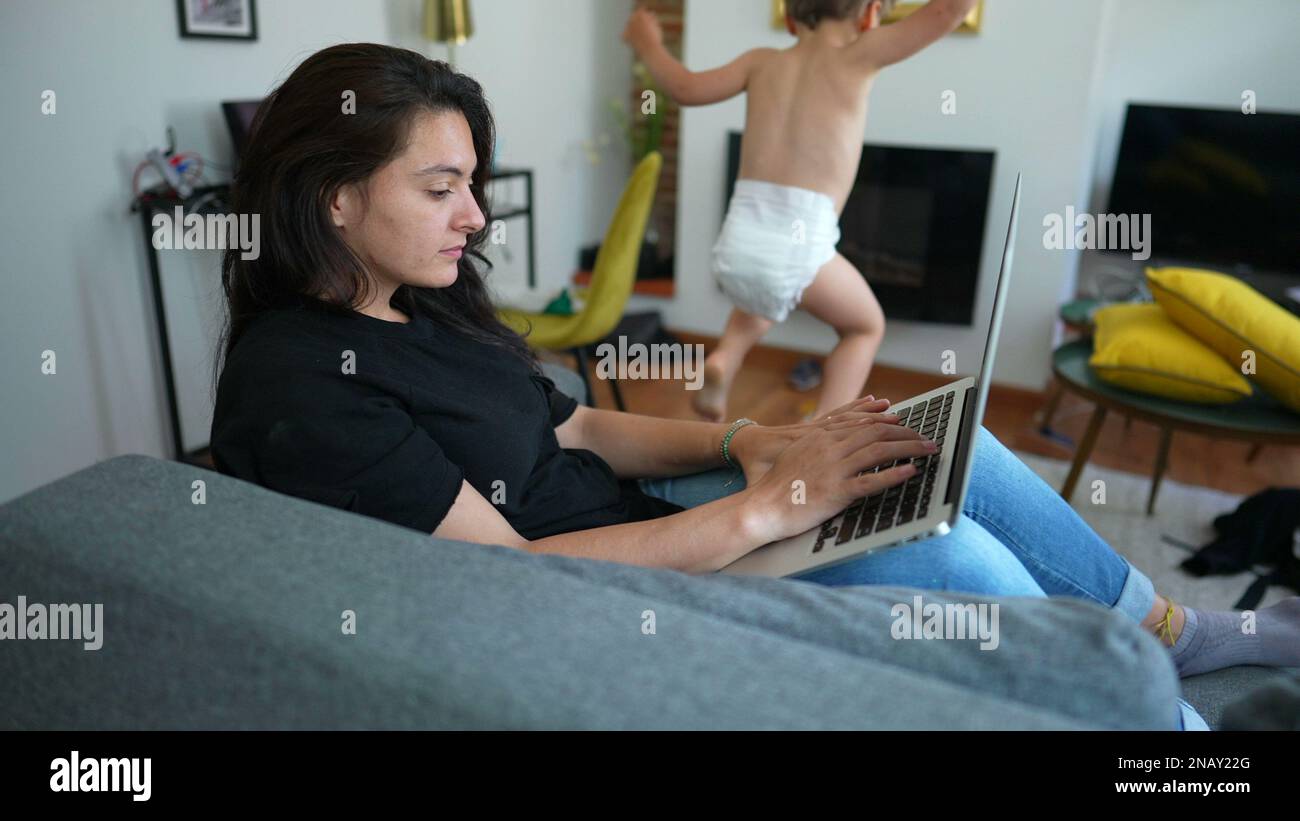 Woman working from home using laptop with child jumping from sofa ...