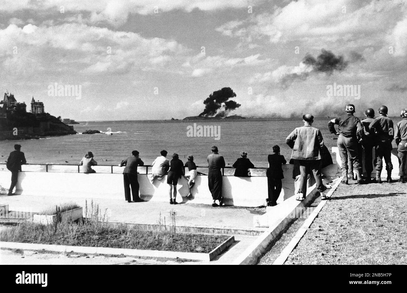 American soldiers and Frenchmen stand along the seawall on the mainland ...