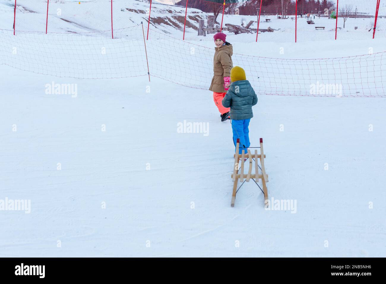 two children pulling the wooden sledge on snow slope in bright clothes ...