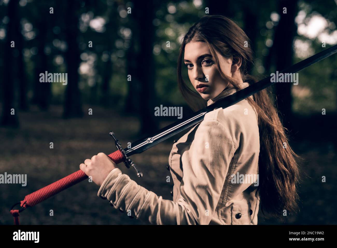 A female in beige costume holding a sword on the shoulder, forest ...