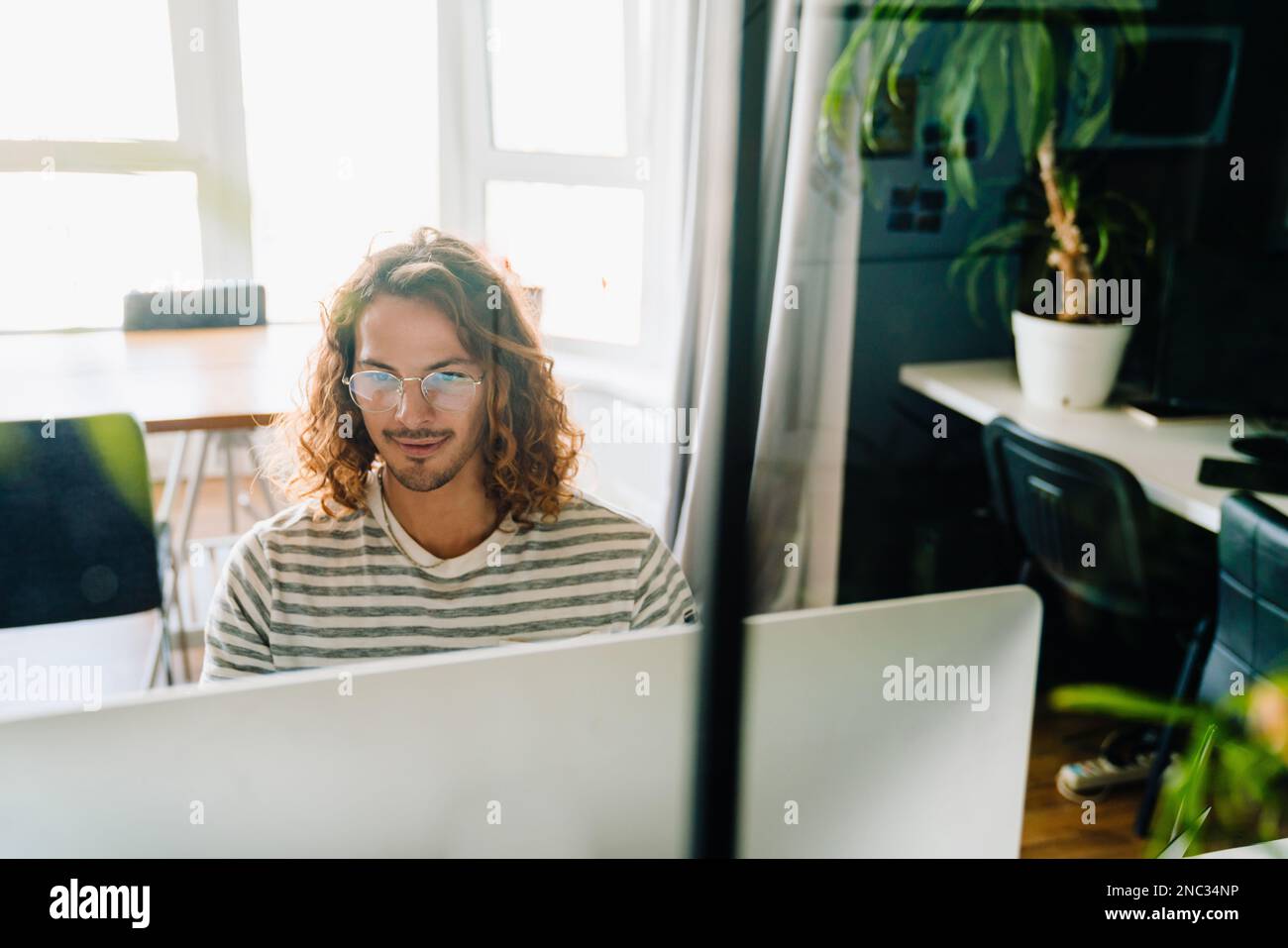 Young curly man using desktop computer while sitting by table in office ...