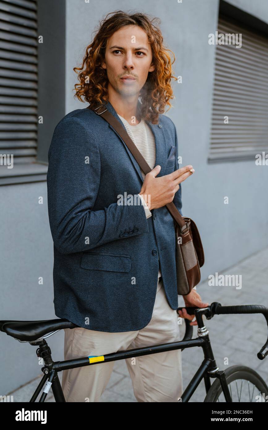 Young ginger long-haired man smoking cigarette while standing with ...