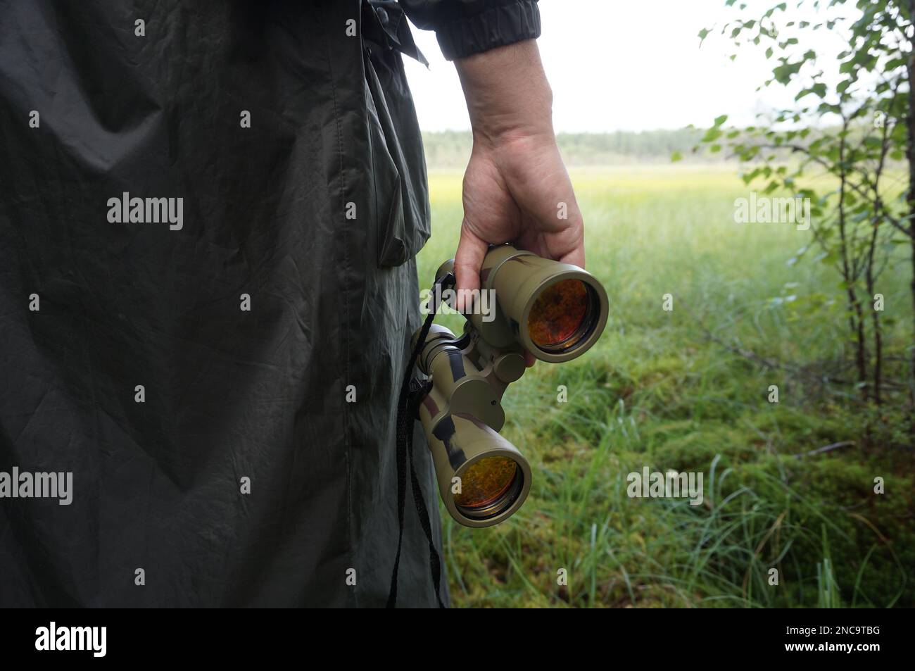 Young man in a raincoat stands near a swamp and holds binoculars in his ...