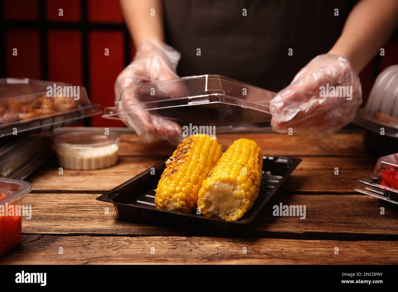 Waiter in gloves closing container with grilled corn cobs at wooden ...