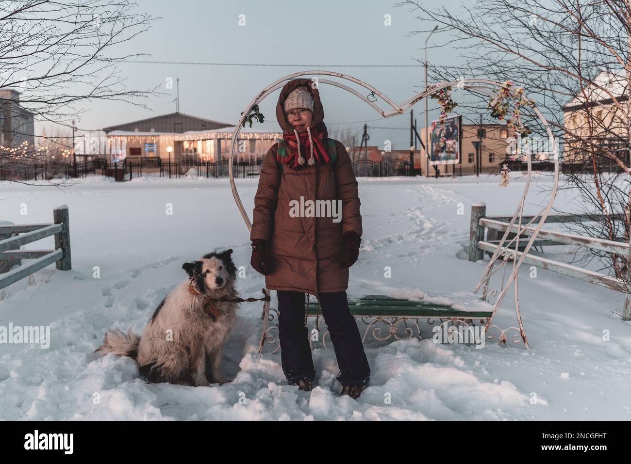 An Asian Yakut girl stands with an old white dog in the snow next to a heart-shaped bench in a ...