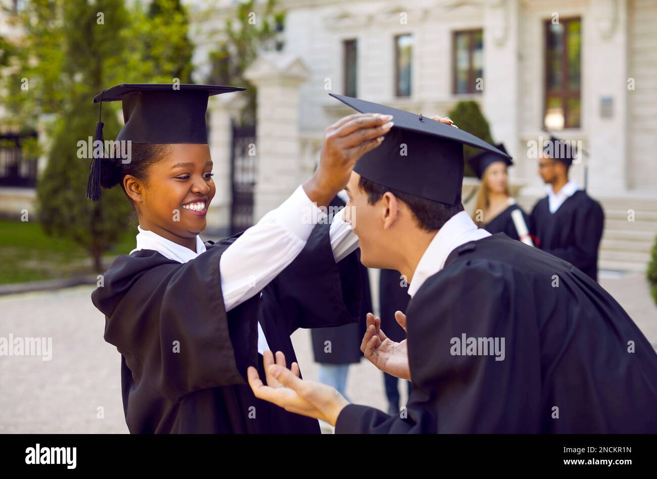 Joyful female student fixing mortarboard to her male classmate before ...