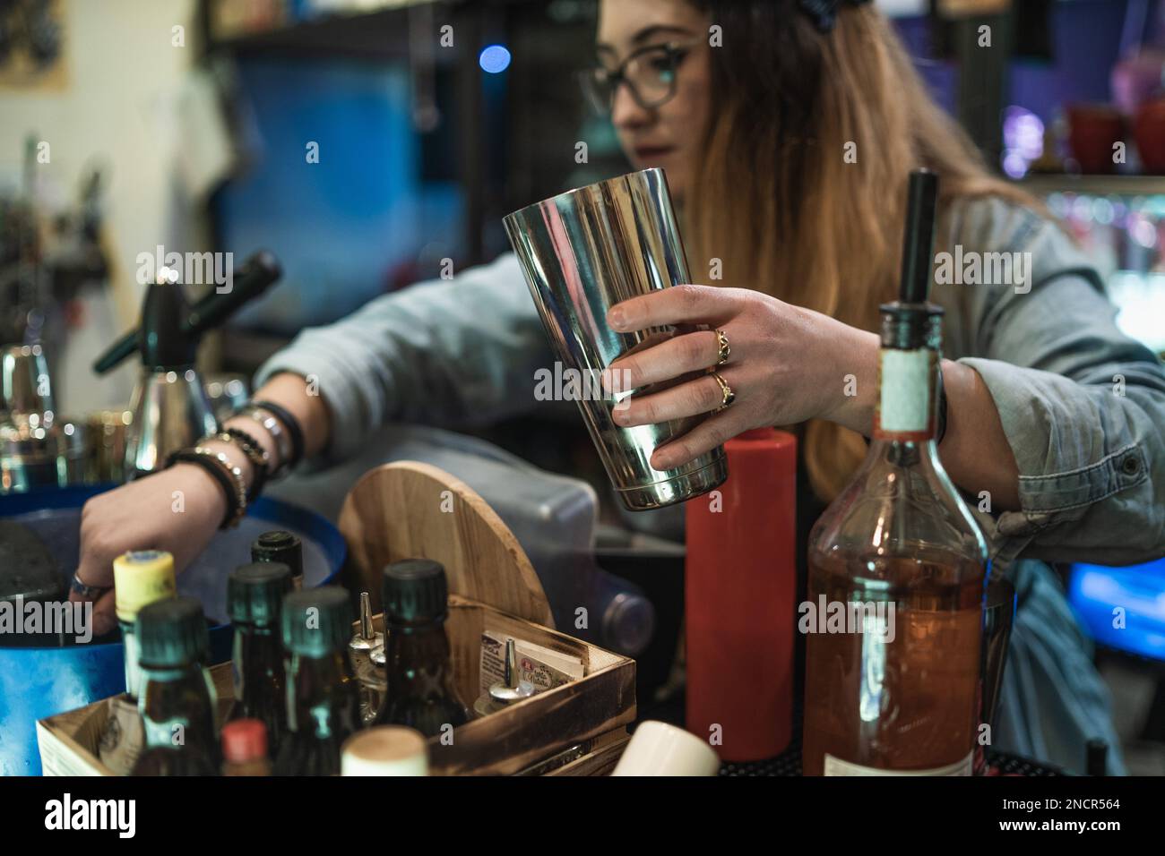 Female bartender making a cocktail behind the bar counter, putting ice in the shaker. Brightly ...