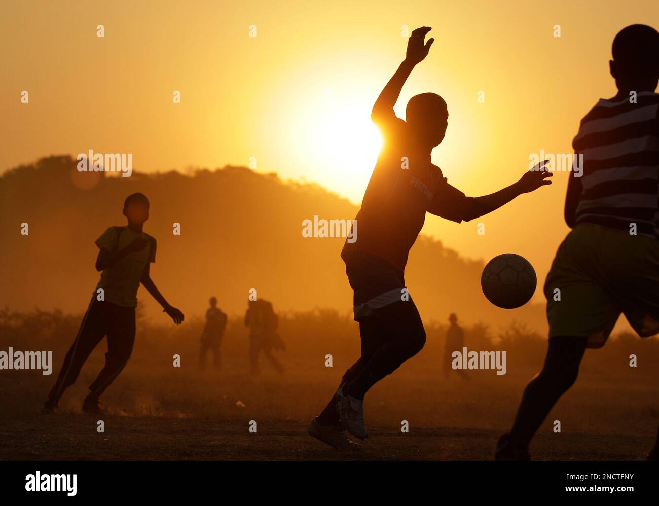 Kids play soccer in a dusty lot as the sun goes down, in a rural ...