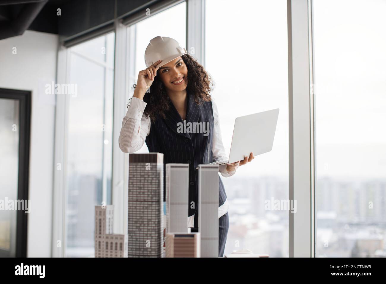 Smiling female architect using laptop inspecting skyscraper model in ...