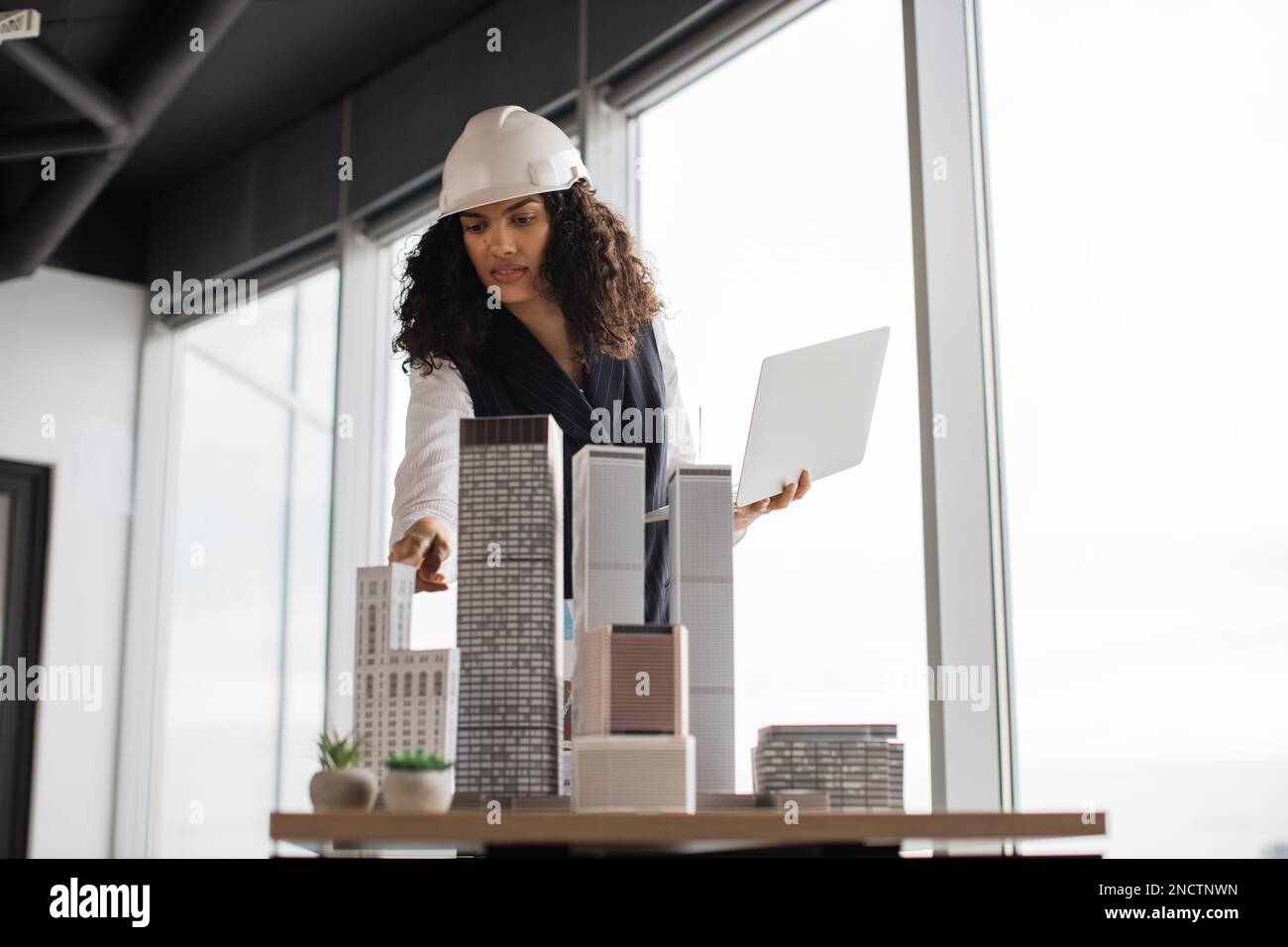 Young female architect using laptop inspecting skyscraper model in ...